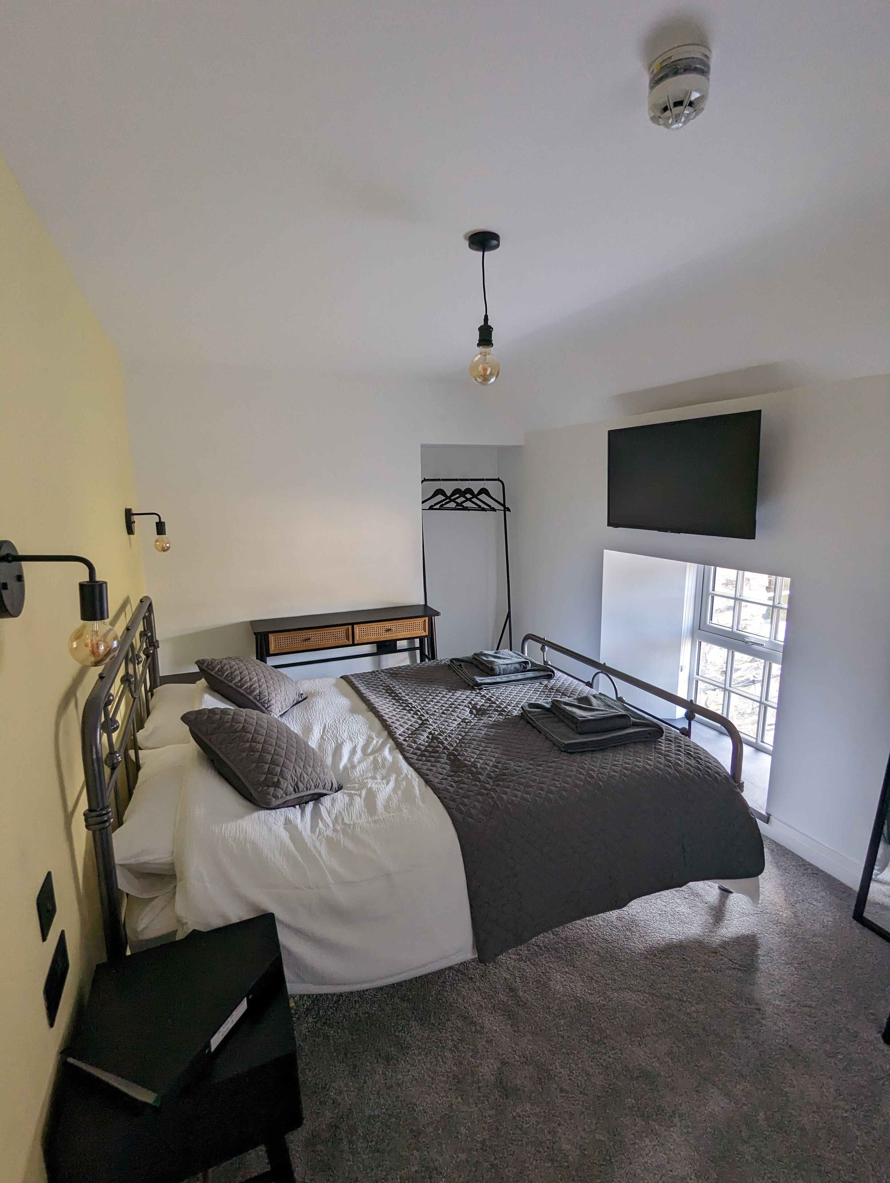 Quarto com cama, mesas de cabeceira, televisão na parede e uma janela.
