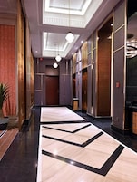 Corredor de hotel com piso estampado em padrão geométrico, balcão de recepção e elevador moderno.