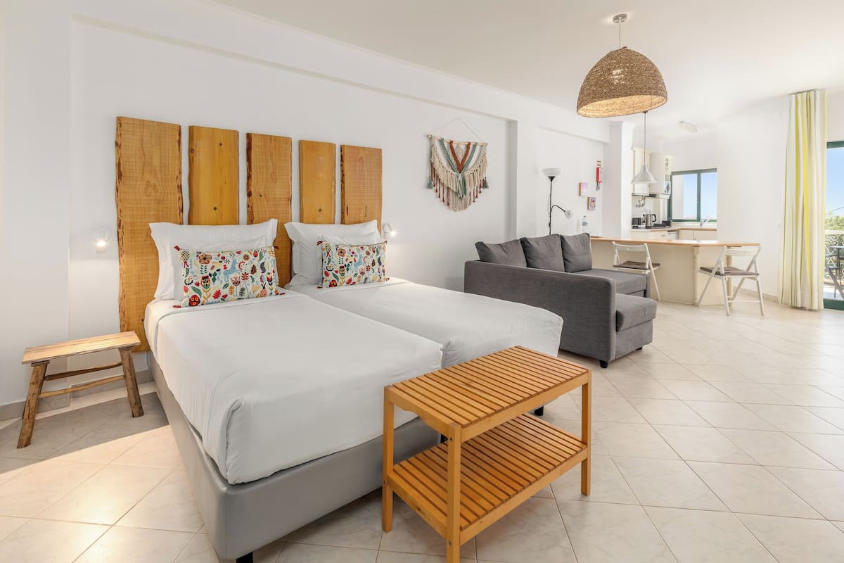 Quarto moderno com uma cama grande, um sofá cinza, uma mesa de cabeceira de madeira e uma cozinha visível através de uma porta aberta.
