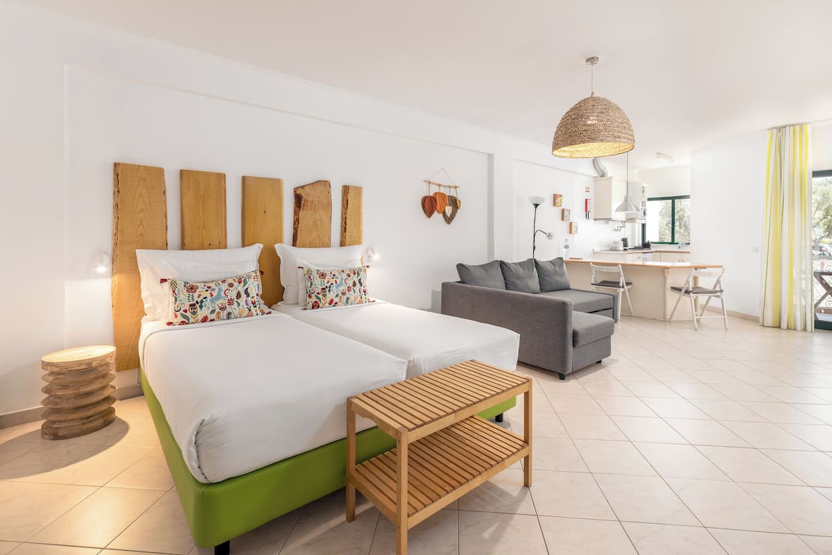 Quarto de hotel moderno com cama verde, sofá cinza, mesa de apoio de madeira e cozinha visível por uma porta aberta.