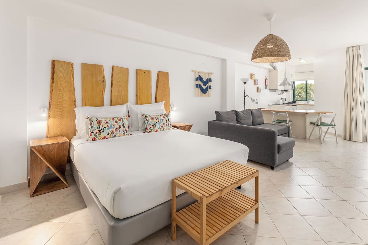Quarto moderno com uma cama grande, um sofá cinza, uma mesinha de cabeceira de madeira e um lustre.
