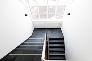 Escadaria moderna com azulejos pretos e corrimão de metal, conduzindo a um espaço iluminado com vista para um pátio revestido de azulejos.