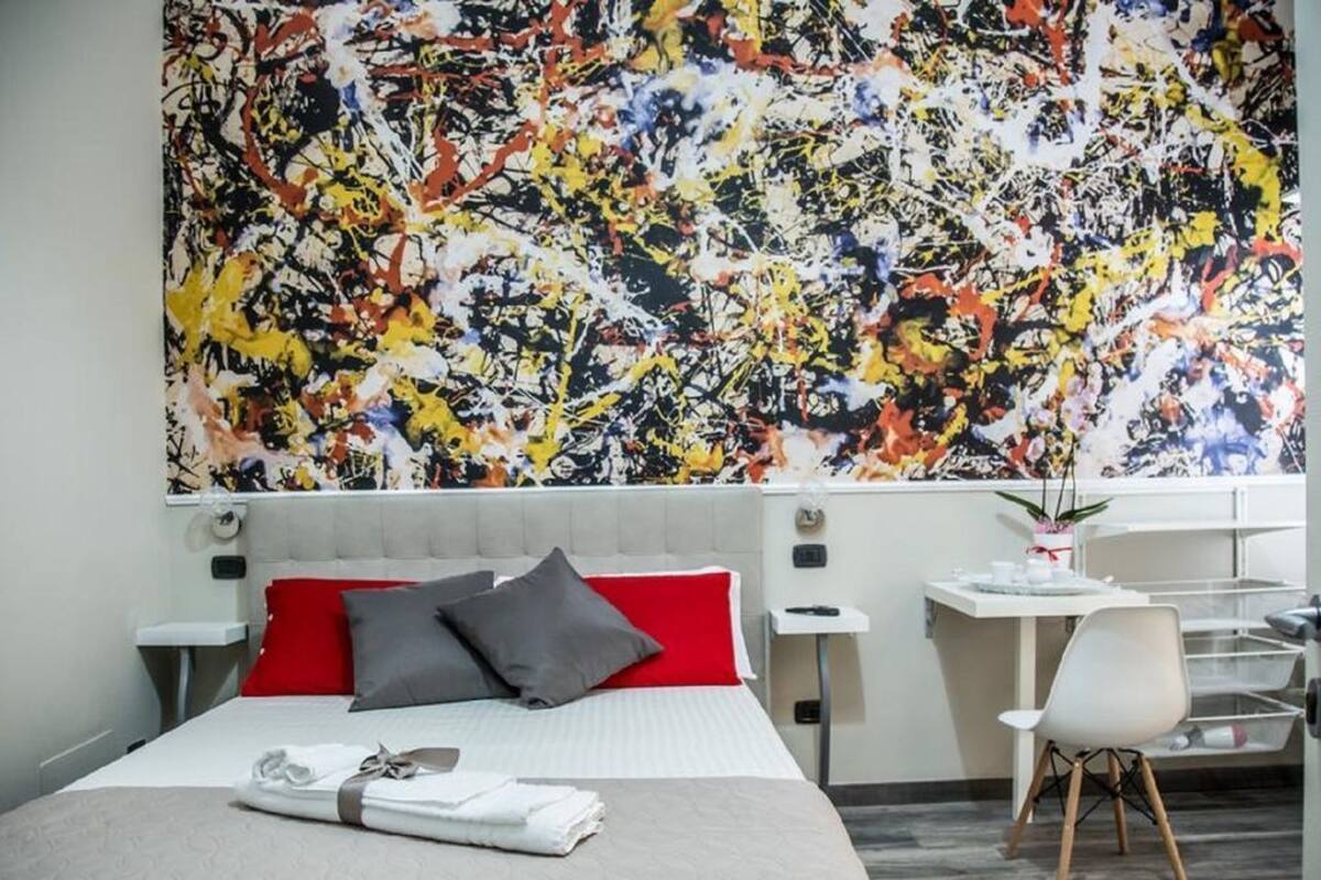 Quarto de hotel moderno com uma grande pintura abstrata na parede, uma cama bem arrumada com almofadas vermelhas e cinzas, e uma pequena escrivaninha com uma cadeira.