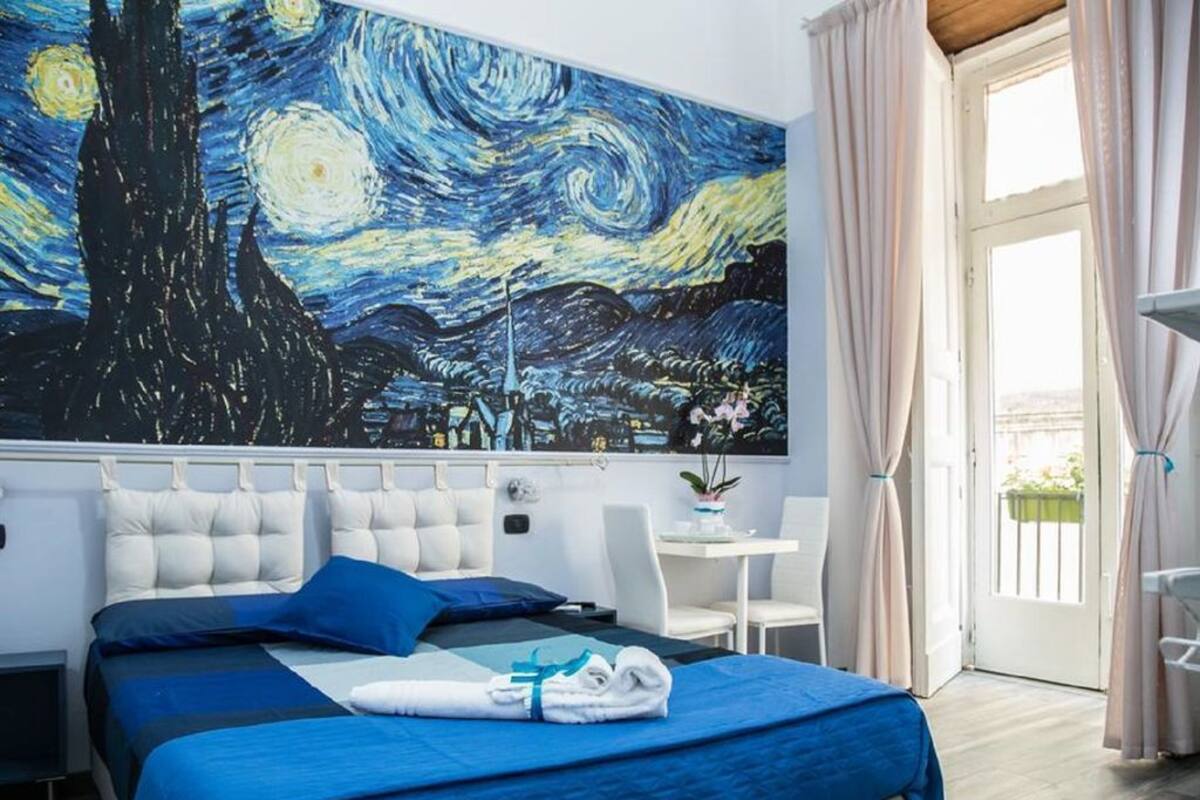 Quarto de hotel com uma grande pintura de uma cena noturna, uma cama com roupa de cama azul, cabeceira branca e uma pequena mesa com uma cadeira.