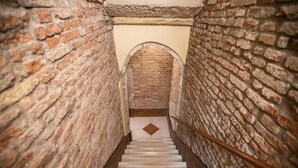 Uma escadaria estreita com paredes de pedra e um corrimão de madeira, dando acesso a uma porta em arco.