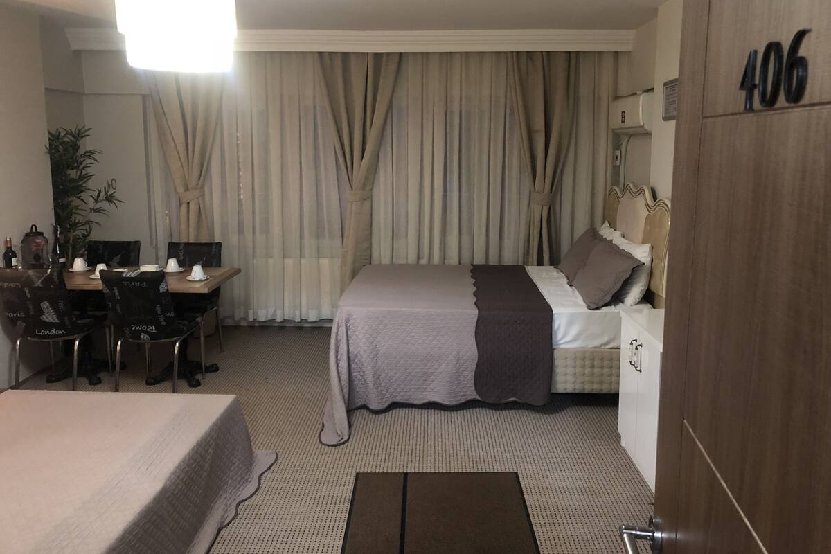 Quarto de hotel com uma cama, uma escrivaninha com cadeira, luminária e cortinas.