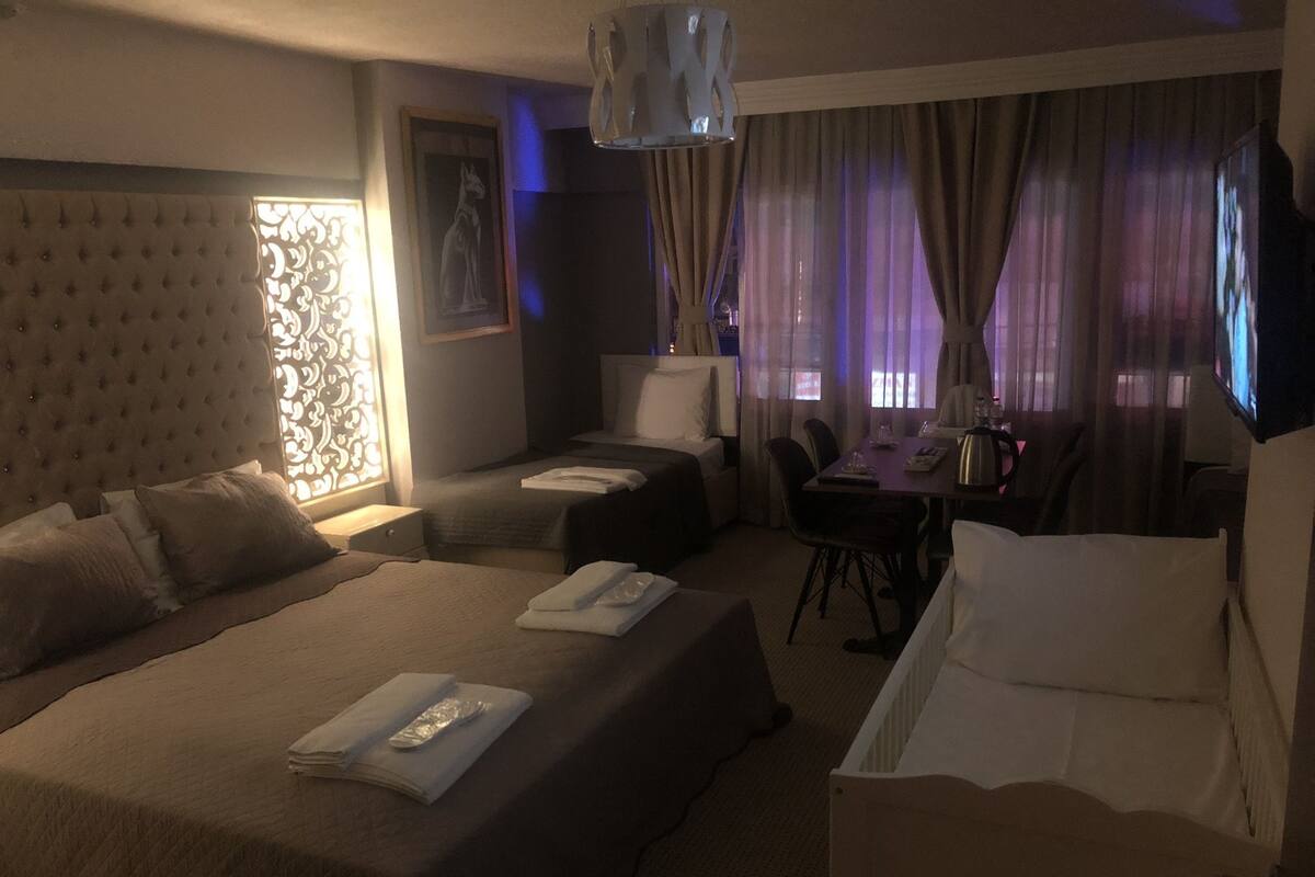 Quarto de hotel com uma cama grande, uma cama menor, uma escrivaninha e uma cadeira. Há uma janela com cortinas, uma luminária e um quadro na parede.