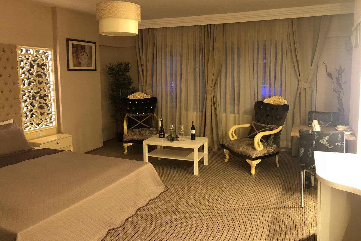 Quarto de hotel com uma cama grande, duas poltronas, uma mesinha de centro com uma garrafa e copos, um painel decorativo e uma janela ampla com cortinas.