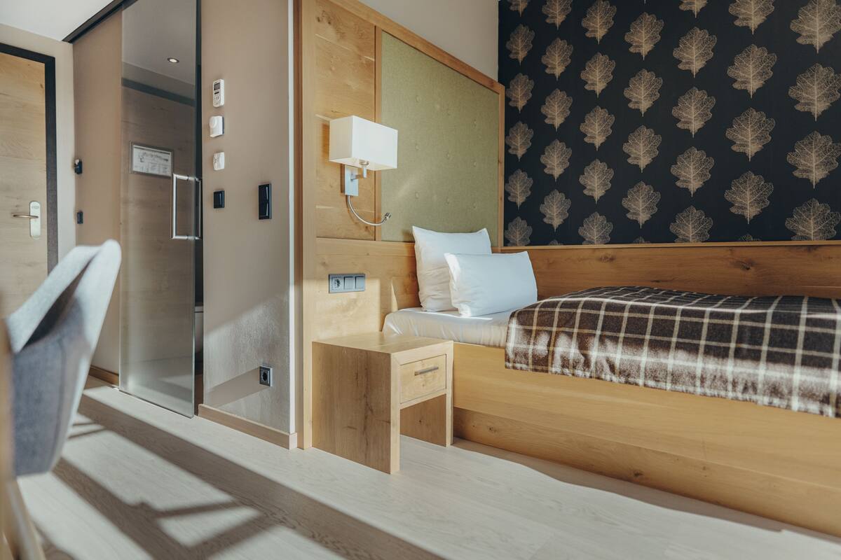 Quarto de hotel moderno com cabeceira de madeira, cama com manta xadrez, mesa de cabeceira com luminária e área de chuveiro.