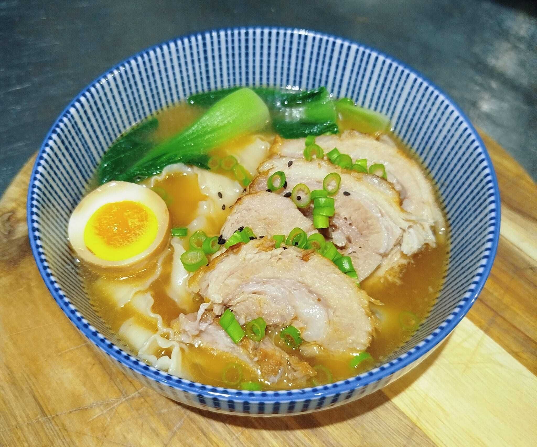 Uma tigela de ramen com fatias de carne de porco, ovo cozido, cebolinha e bok choy numa tigela com borda azul.