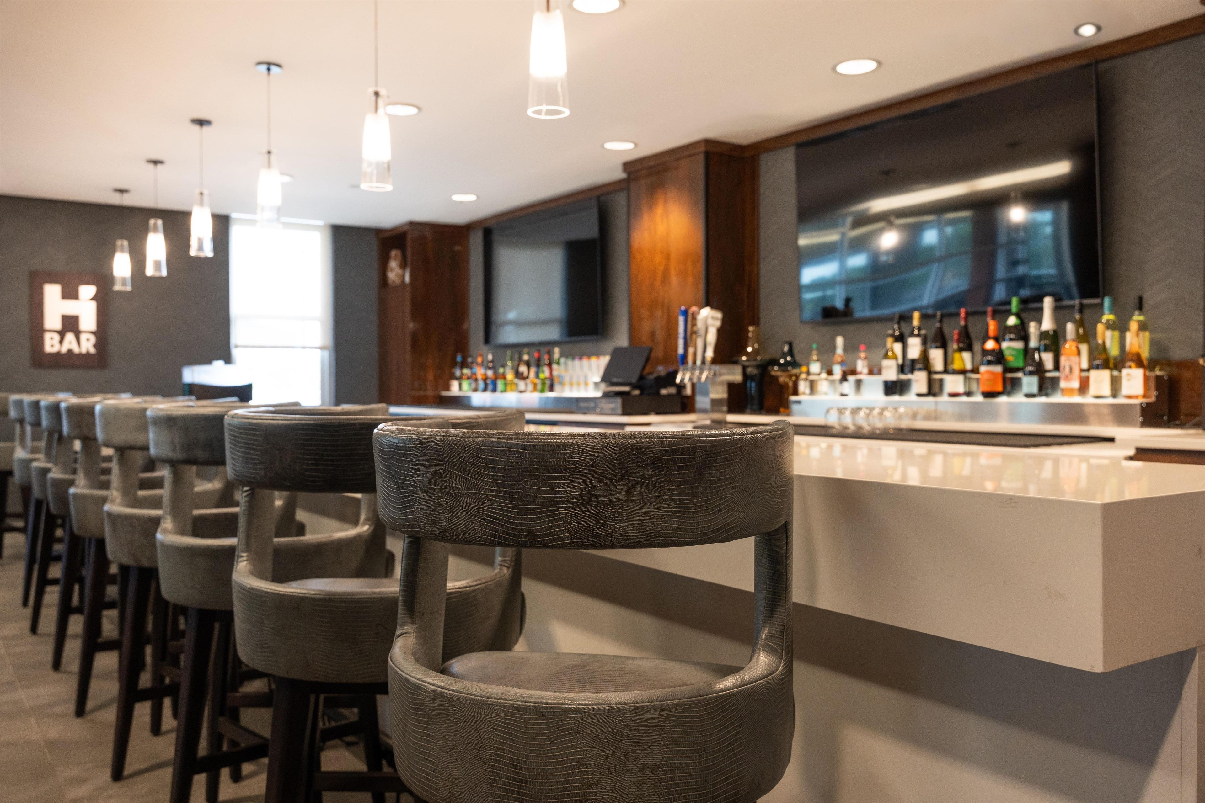 Uma área de bar com vários bancos altos e um balcão exibindo diversos tipos de bebidas.