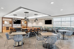 Uma sala de jantar moderna com várias mesas e cadeiras, uma cozinha ao fundo e grandes janelas.