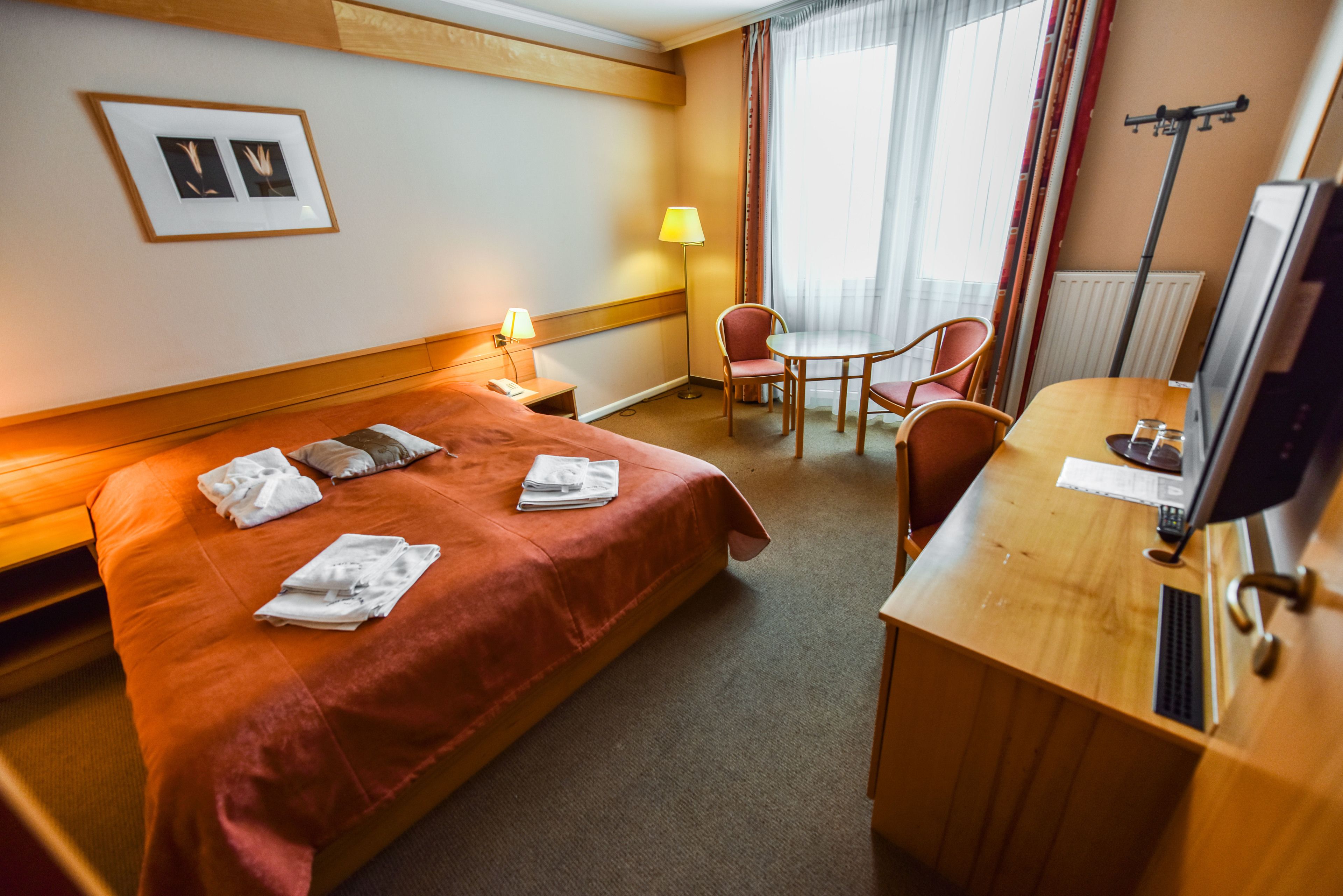 Quarto de hotel com cama, escrivaninha, cadeira, abajur e televisão.
