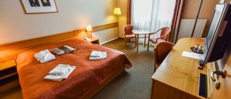 Quarto de hotel com cama, escrivaninha, cadeira, abajur e televisão.