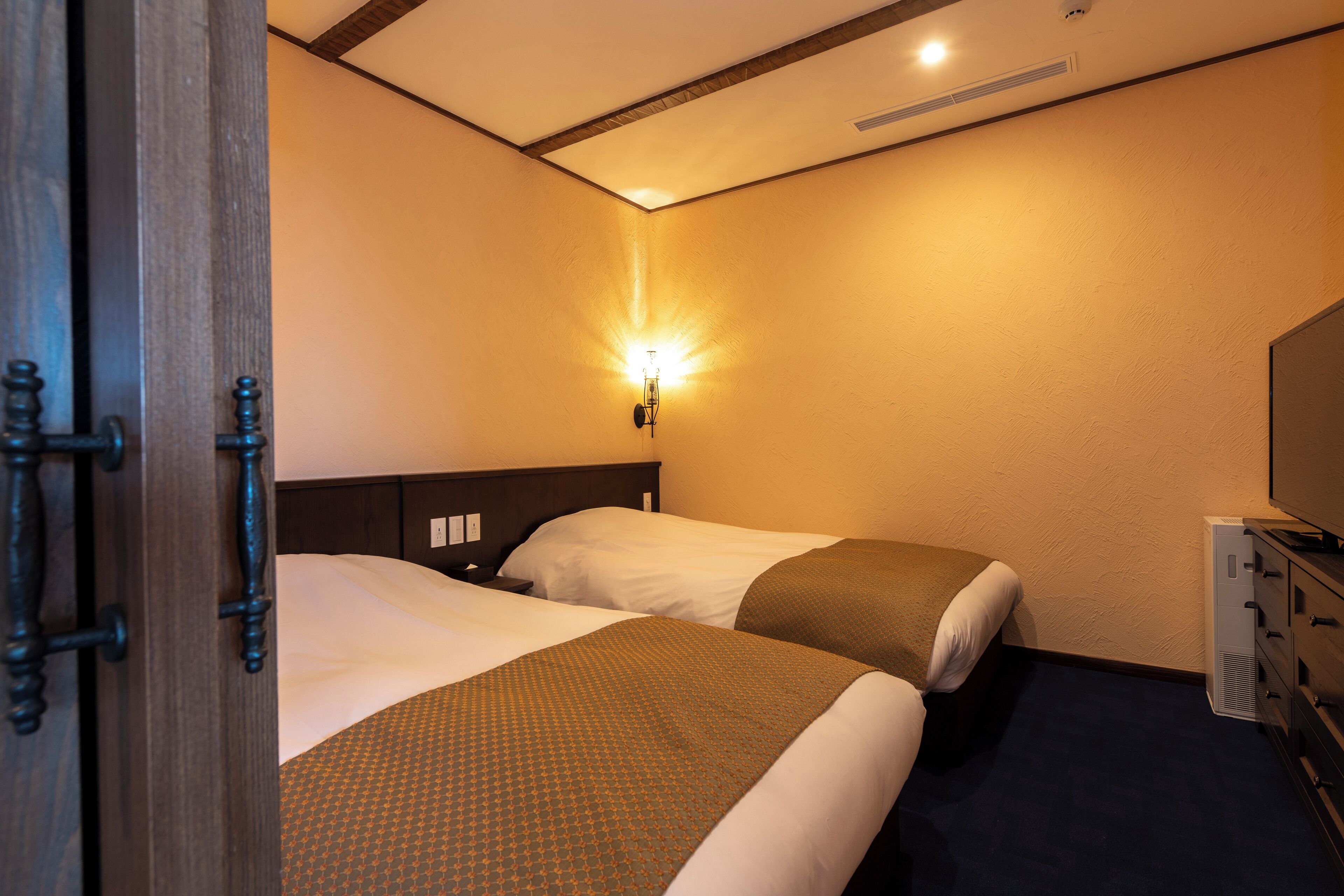 Quarto de hotel com duas camas, uma mesinha de cabeceira, um penteado e uma luminária fixada na parede.