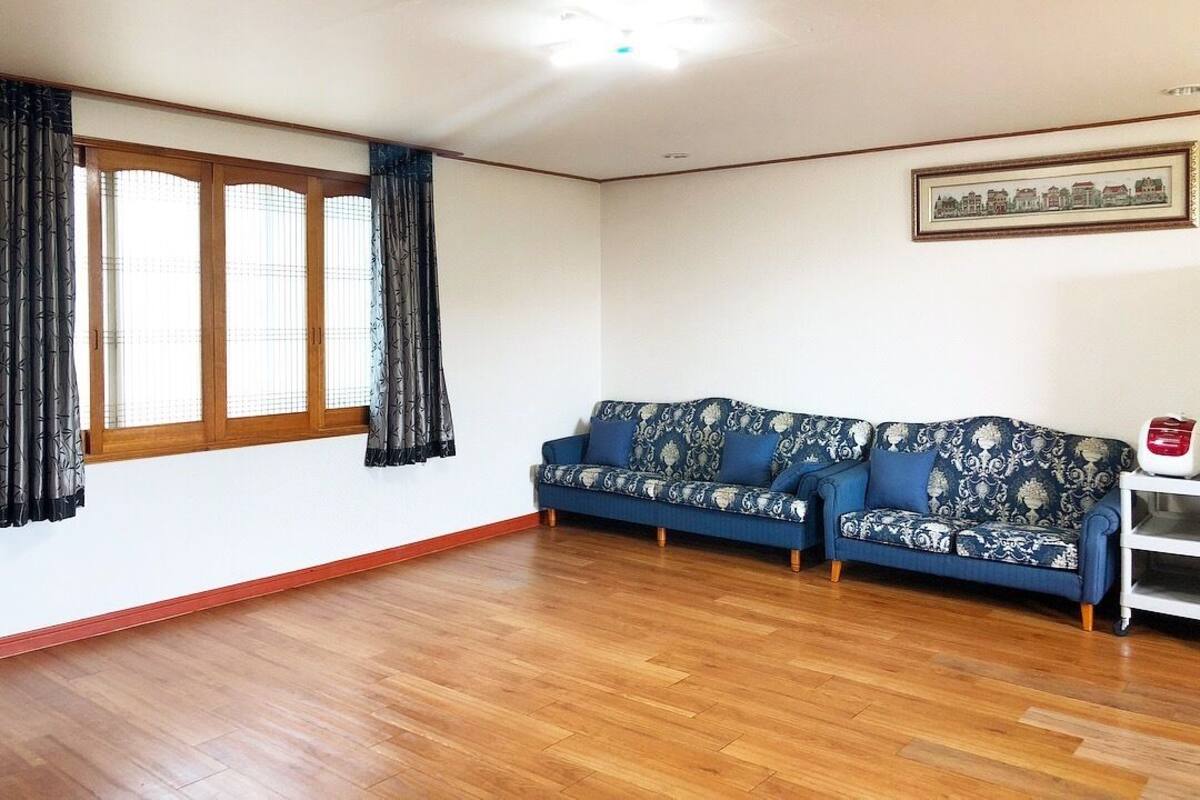 Uma sala de estar com um sofá azul estampado, piso de madeira, uma janela com cortinas e um quadro na parede.