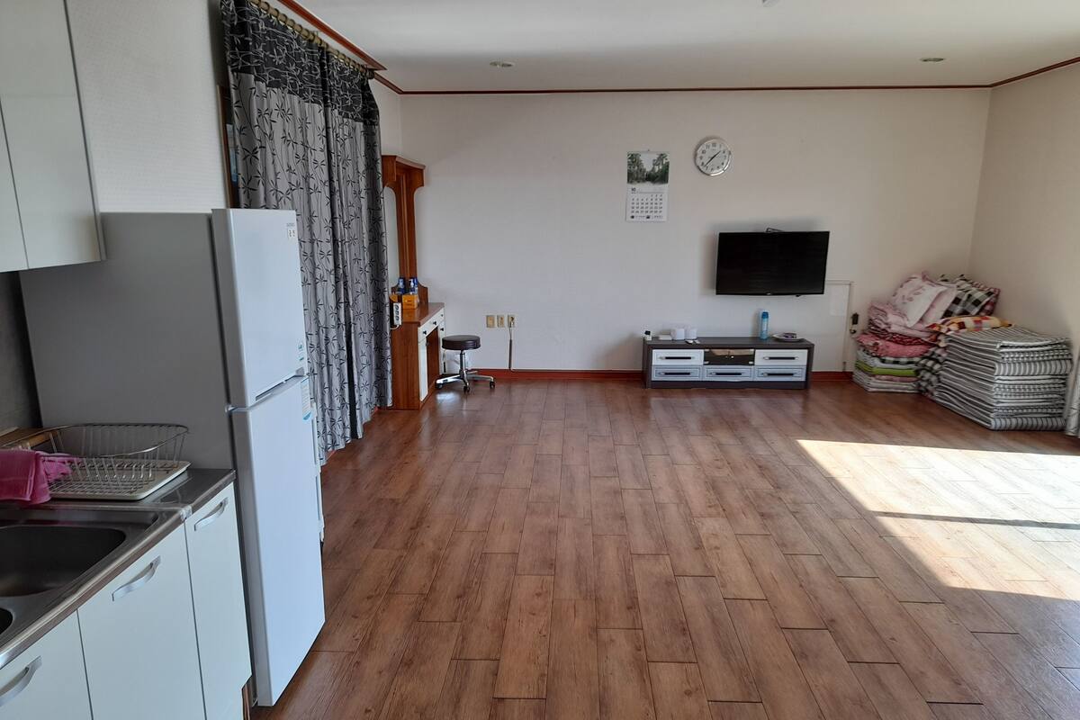 Uma sala de estar espaçosa com piso de madeira, cozinha equipada com geladeira, mesa de jantar e televisão na parede.