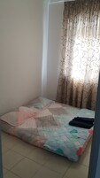 Uma cama pequena e bem arrumada, com colcha acolchoada e almofadas, uma janela com cortinas estampadas e piso de azulejos.