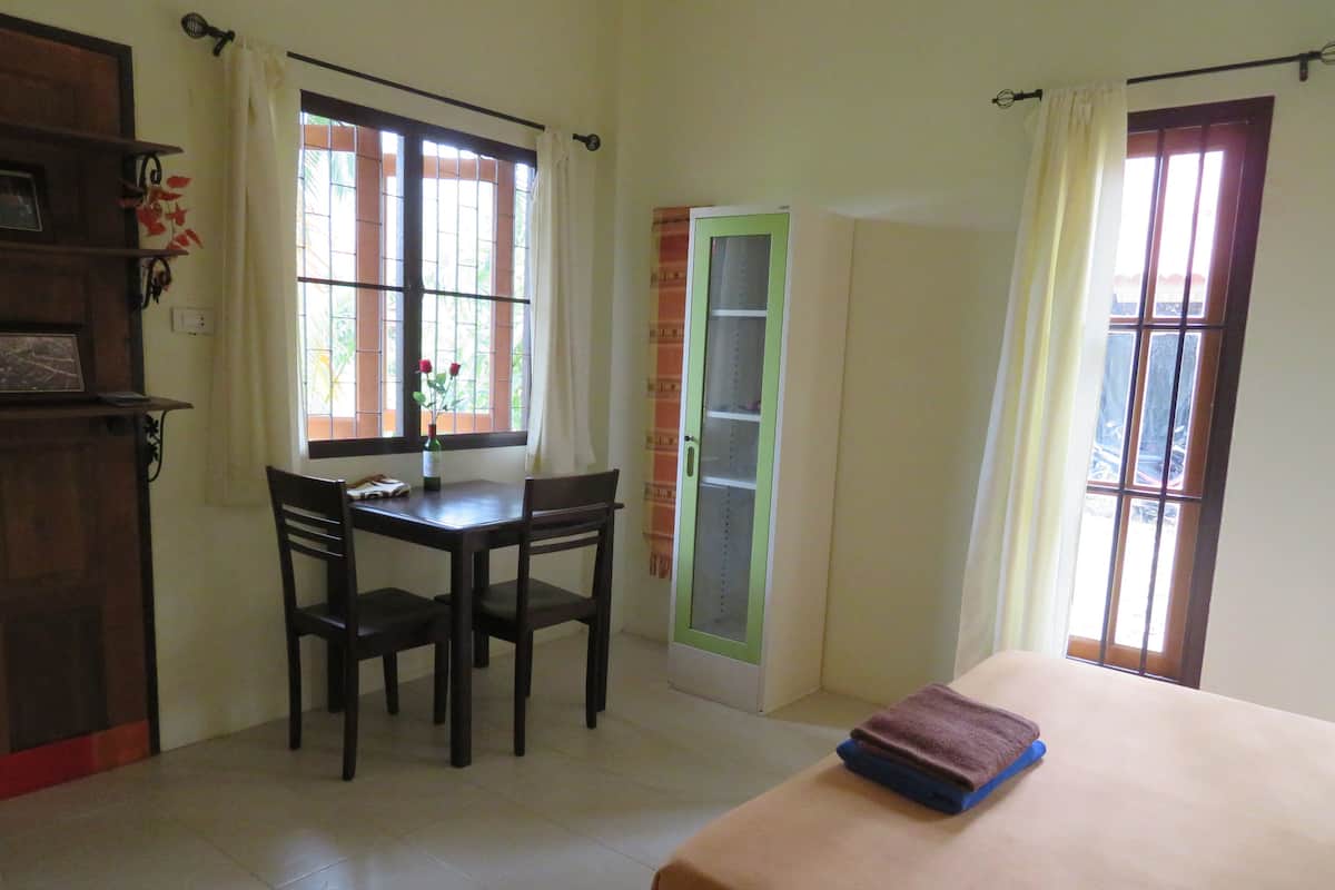 Quarto com cama, escrivaninha e duas cadeiras, janela com cortinas e porta com moldura verde.