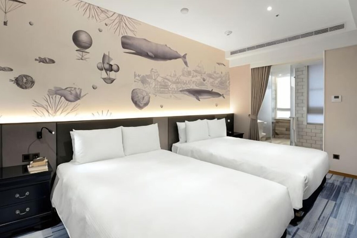 Quarto de hotel com duas camas, mesa de cabeceira, painel decorativo na parede e porta de correr para uma varanda.