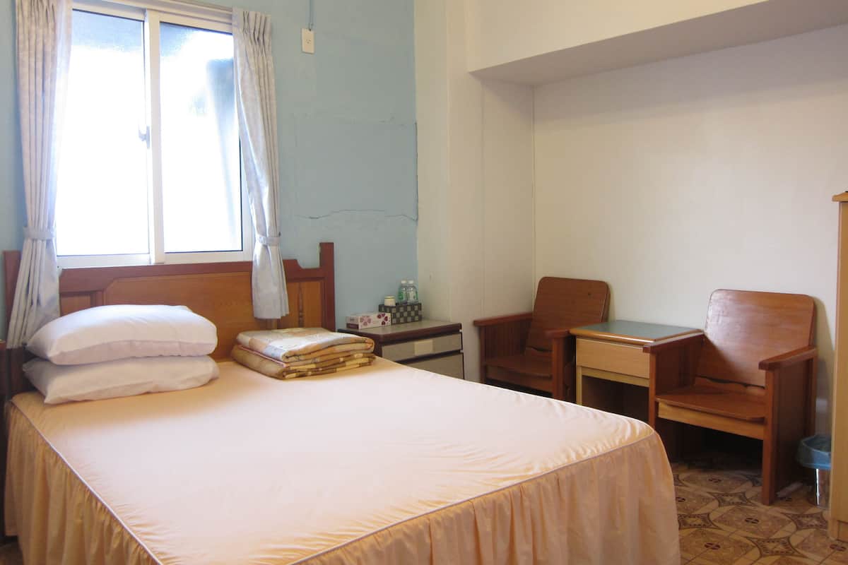 Quarto simples com uma cama, duas cadeiras, uma mesinha e uma janela com cortinas.