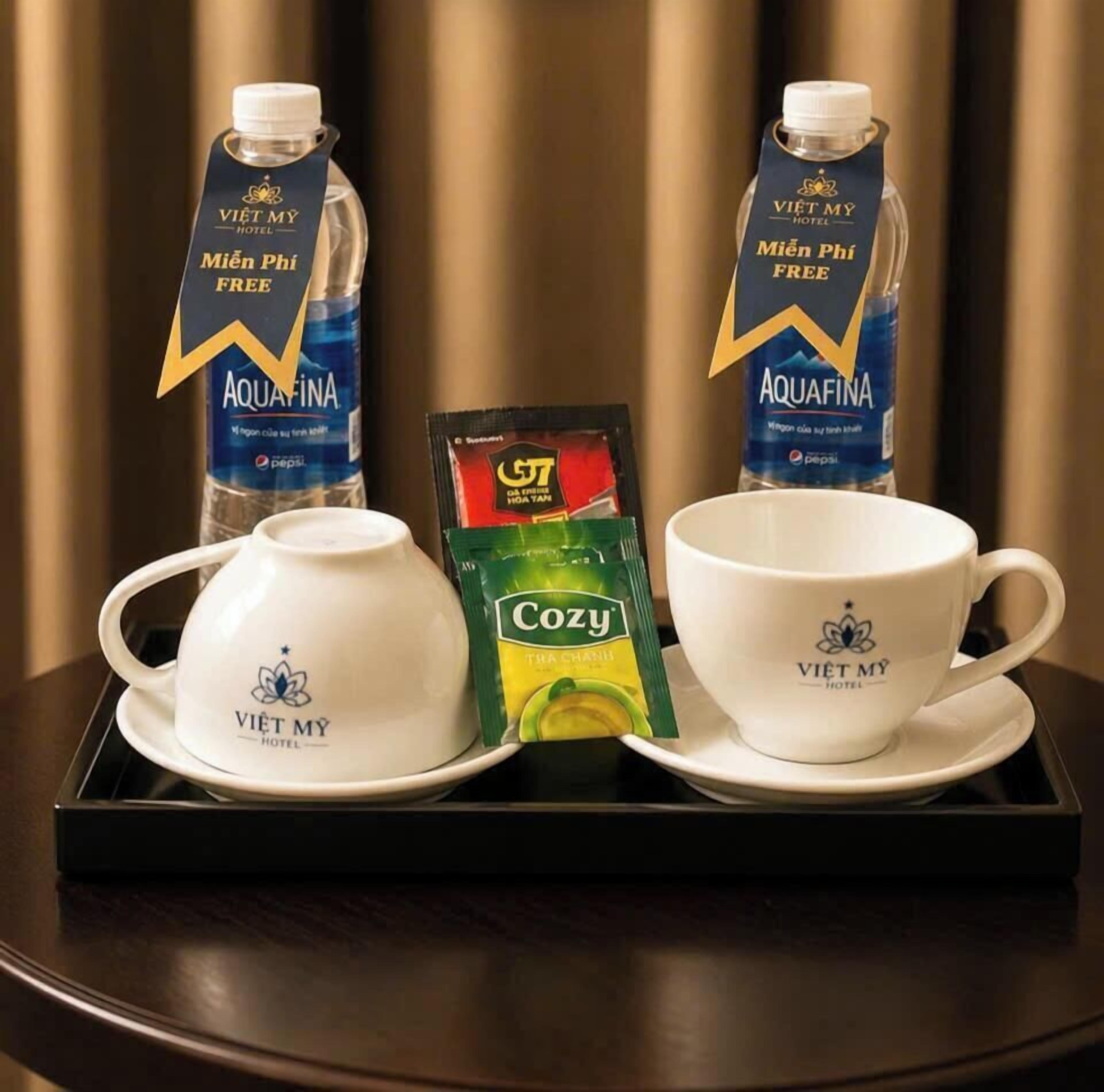 Uma bandeja com duas garrafas de água Aquafina, um sachê de chá Cozy e duas xícaras brancas com o logotipo do Hotel Viet My.