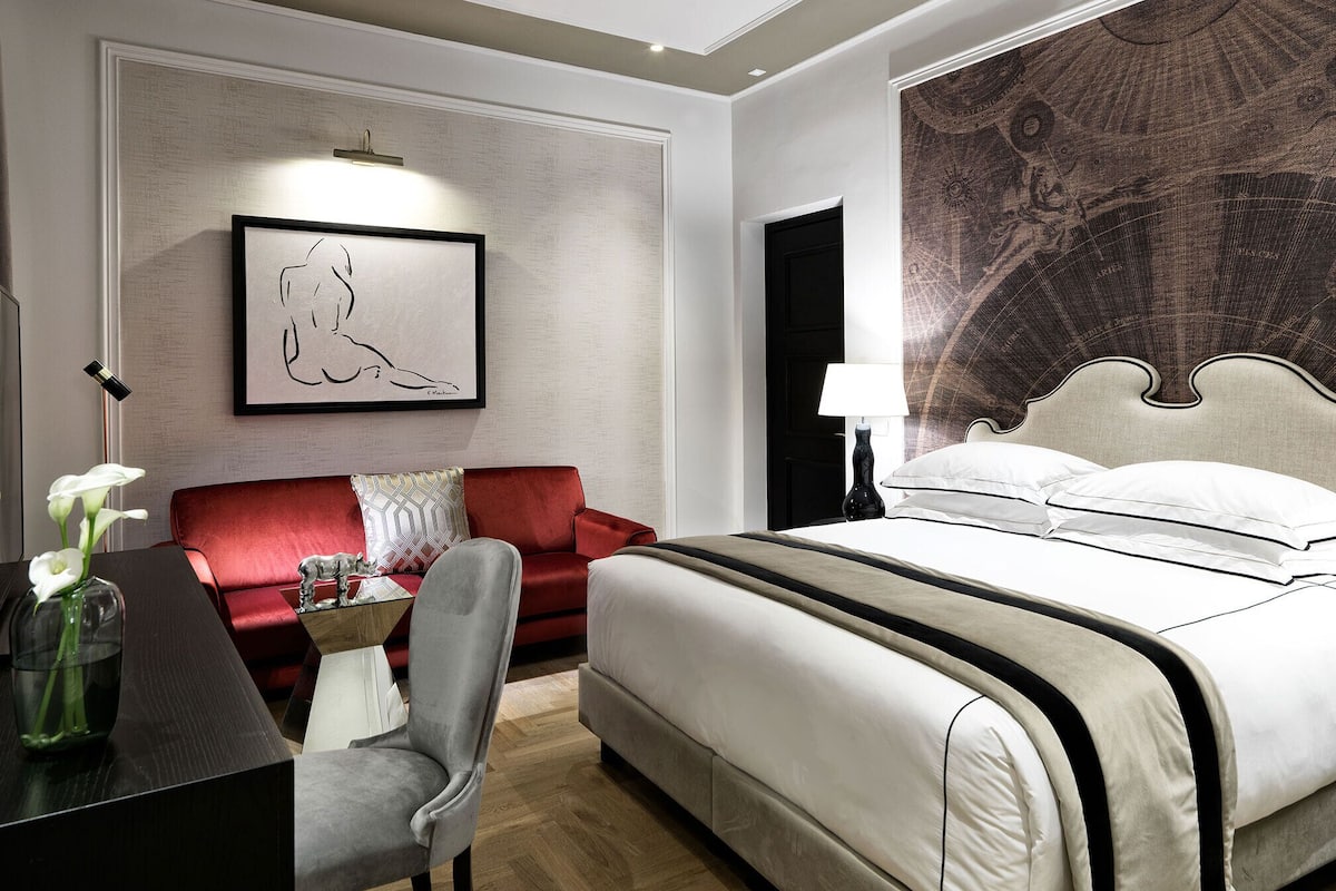 Quarto de hotel com cama, sofá vermelho, escrivaninha com cadeira e um grande tapeçaria na parede.