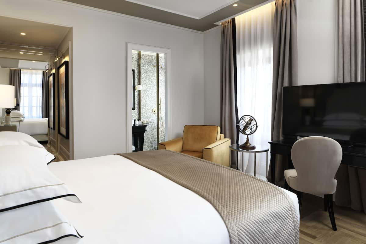 Quarto de hotel com uma cama grande, uma escrivaninha com televisão, uma cadeira e uma mesa pequena com um objeto decorativo.
