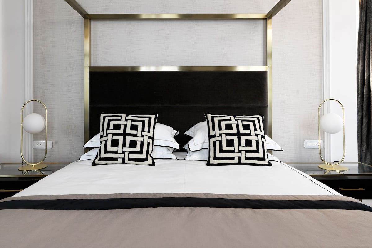 Quarto de hotel moderno com uma cama grande, duas mesinhas de cabeceira e um painel de cabeceira.