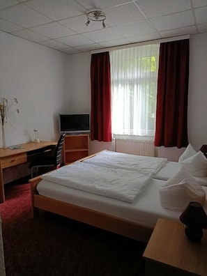 Quarto de hotel com cama, escrivaninha, televisão e janela com cortinas.