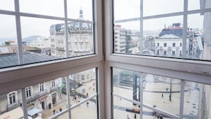 Visão pela janela com padrão de grade, mostrando uma praça com pessoas, edifícios históricos e uma estrutura moderna com cúpula.