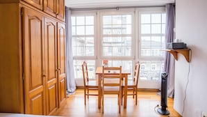 Quarto com armários de madeira, mesa de jantar com cadeiras, aquecedor portátil e grandes janelas com vista para a cidade.