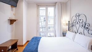 Um quarto com cama, televisão na parede, janela com vista para edifícios, mesa de cabeceira e criado-mudo.