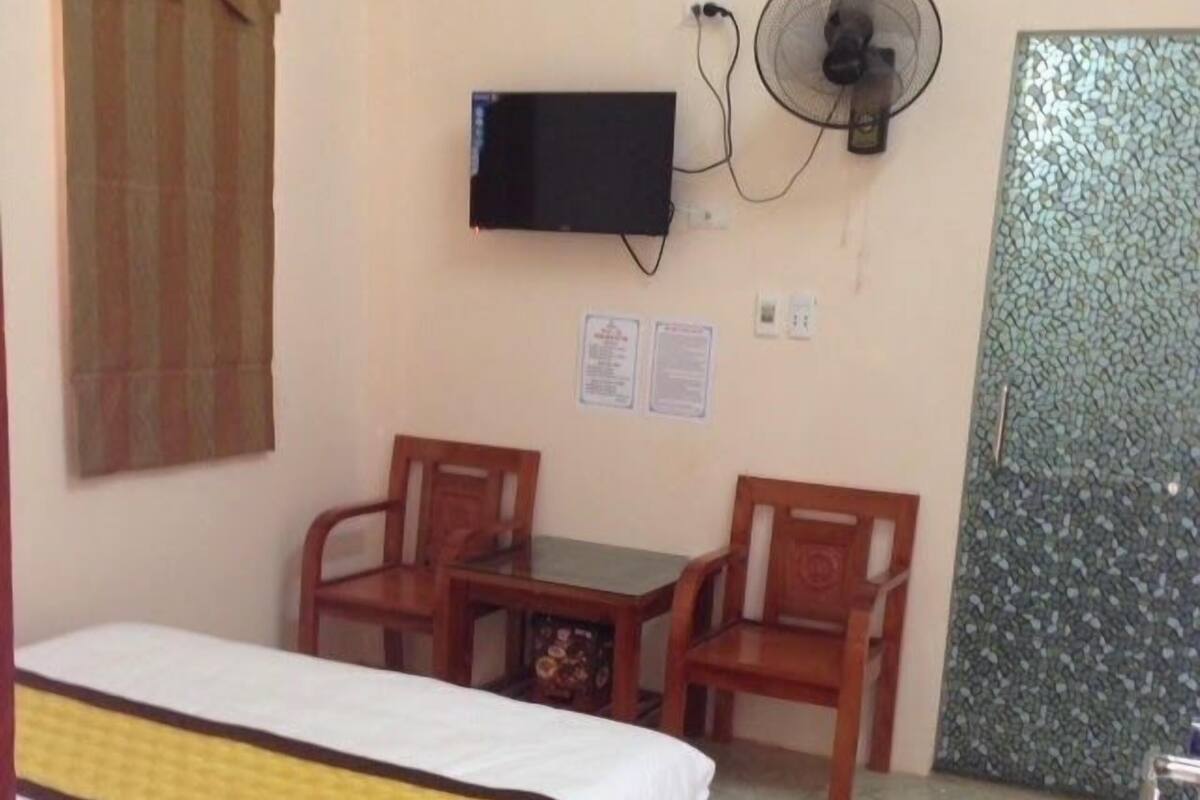 Quarto de hotel com uma cama, uma mesinha, duas cadeiras, uma televisão e um ventilador fixado na parede.