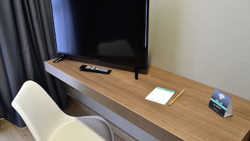 Quarto de hotel com televisão de tela plana sobre uma escrivaninha de madeira, controle remoto, bloco de notas, lápis e sinal de Wi-Fi.