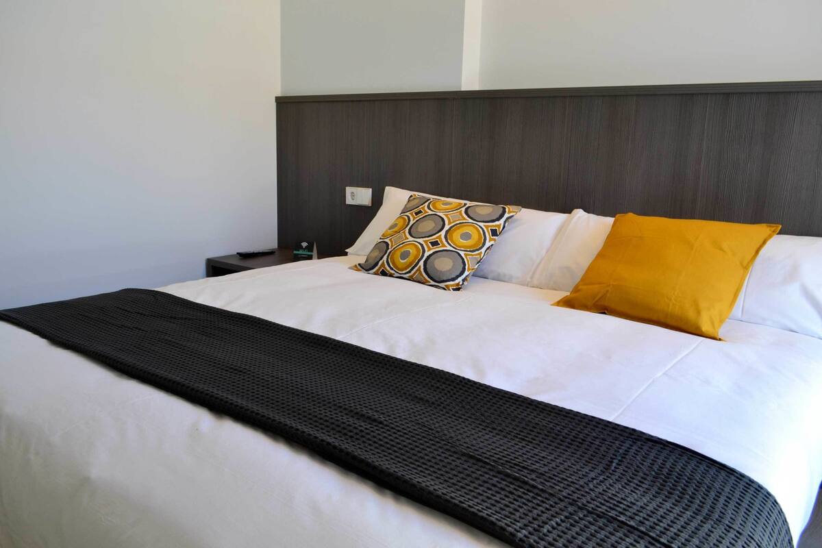 Cama bem arrumada com roupa de cama branca, manta preta e duas almofadas decorativas.