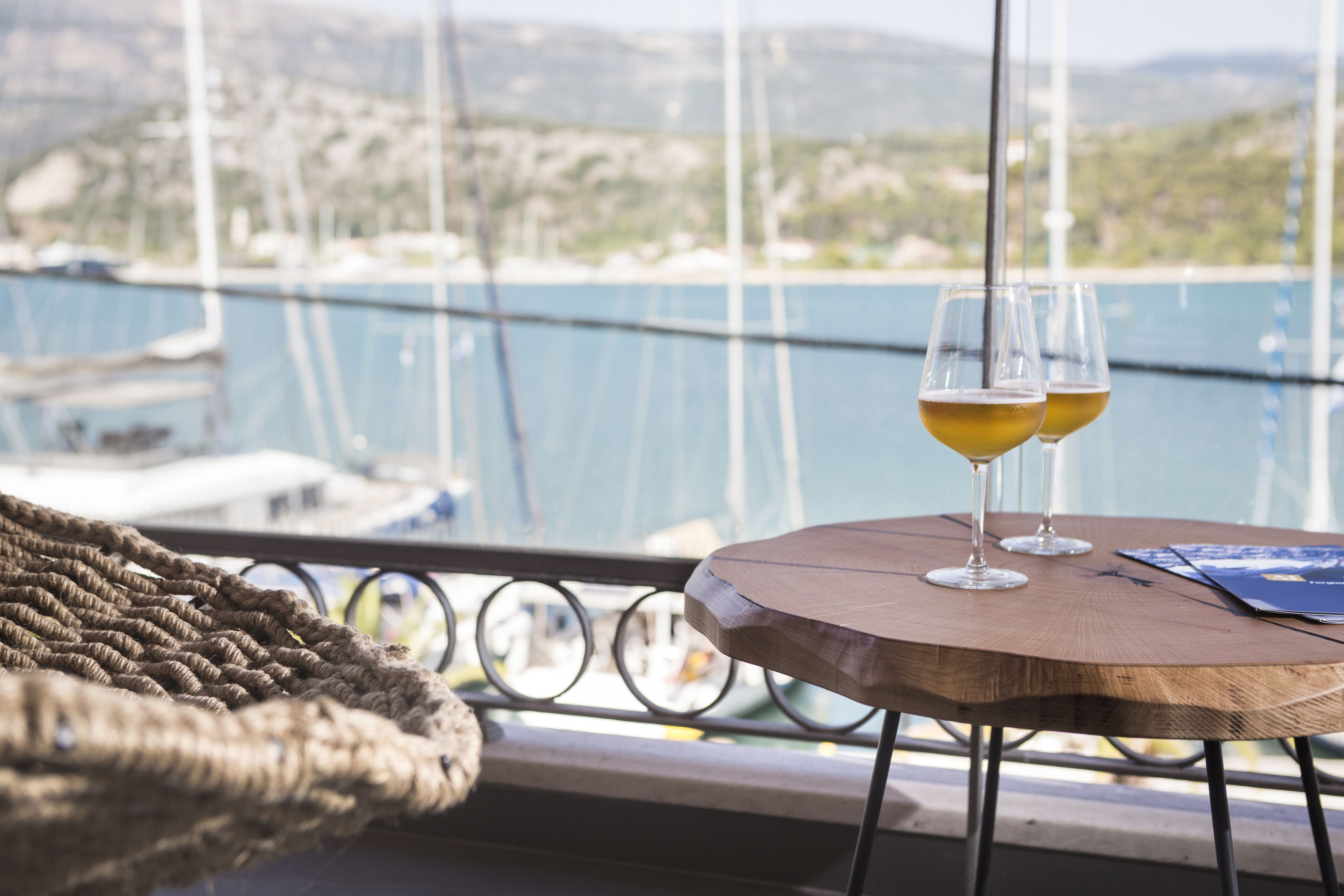 Mesa de madeira redonda com duas taças de vinho numa varanda com vista para uma marina, barcos e paisagem montanhosa.