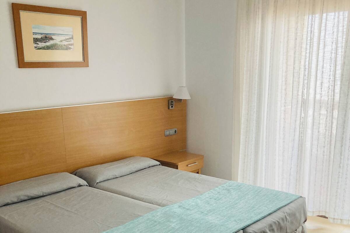 Uma cama de casal com colcha cinza e verde-água, uma cabeceira de madeira, uma mesa de cabeceira com abajur e um quadro emoldurado na parede.