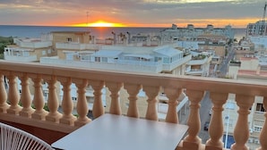 Varanda com mesa e duas cadeiras, vista para a paisagem urbana ao pôr do sol.
