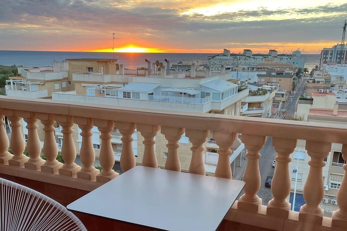 Varanda com mesa e duas cadeiras, vista para a paisagem urbana ao pôr do sol.