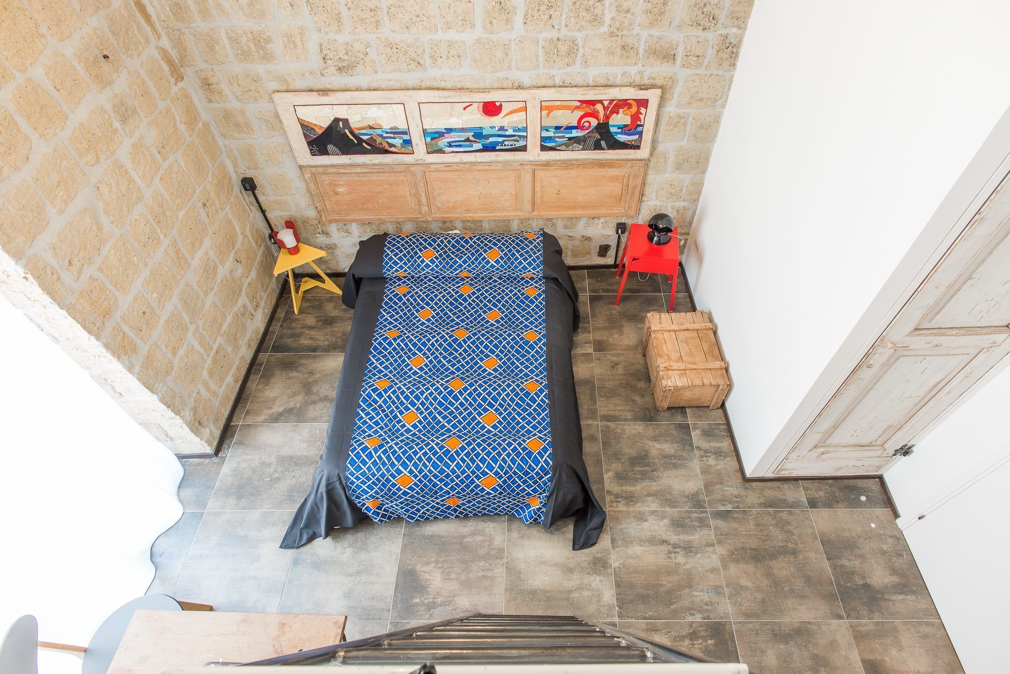 Quarto pequeno com uma cama, uma mesinha e uma cadeira. Uma parede exibe um padrão de azulejos e há um banco de madeira.