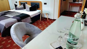 Quarto de hotel com uma cama, uma pequena mesa com uma garrafa e petiscos, uma cadeira e um tapete vermelho.