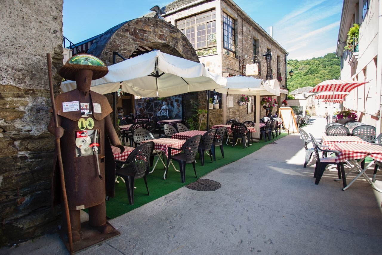 Um café ao ar livre com mesas e cadeiras, uma estátua rústica e um edifício com fachada de pedra.