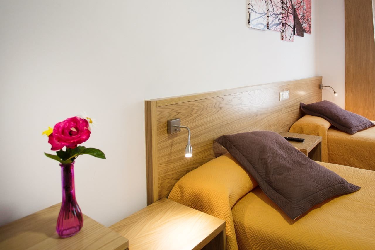Quarto de hotel com cabeceira de madeira, mesa de cabeceira com abajur, um vaso com flor e cama com roupa de cama amarela e travesseiro marrom.