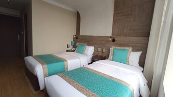 Quarto de hotel com duas camas, cabeceira de madeira, mesas de cabeceira com luminárias e uma porta à esquerda.