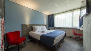 Quarto de hotel com uma cama, uma cadeira vermelha, uma escrivaninha e uma janela com cortinas.