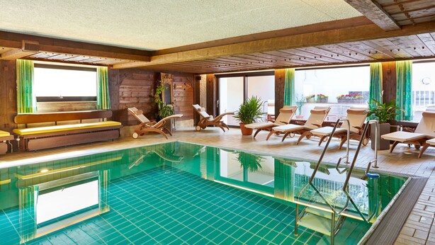 Piscina interior com água cristalina, espreguiçadeiras de madeira e plantas em vasos.