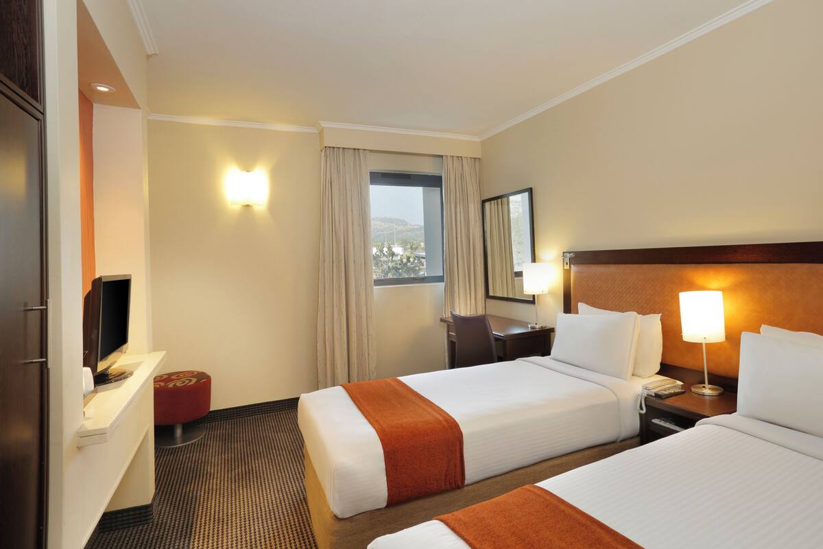 Quarto de hotel com duas camas, uma escrivaninha, televisão, janela com cortinas e vista para o exterior.