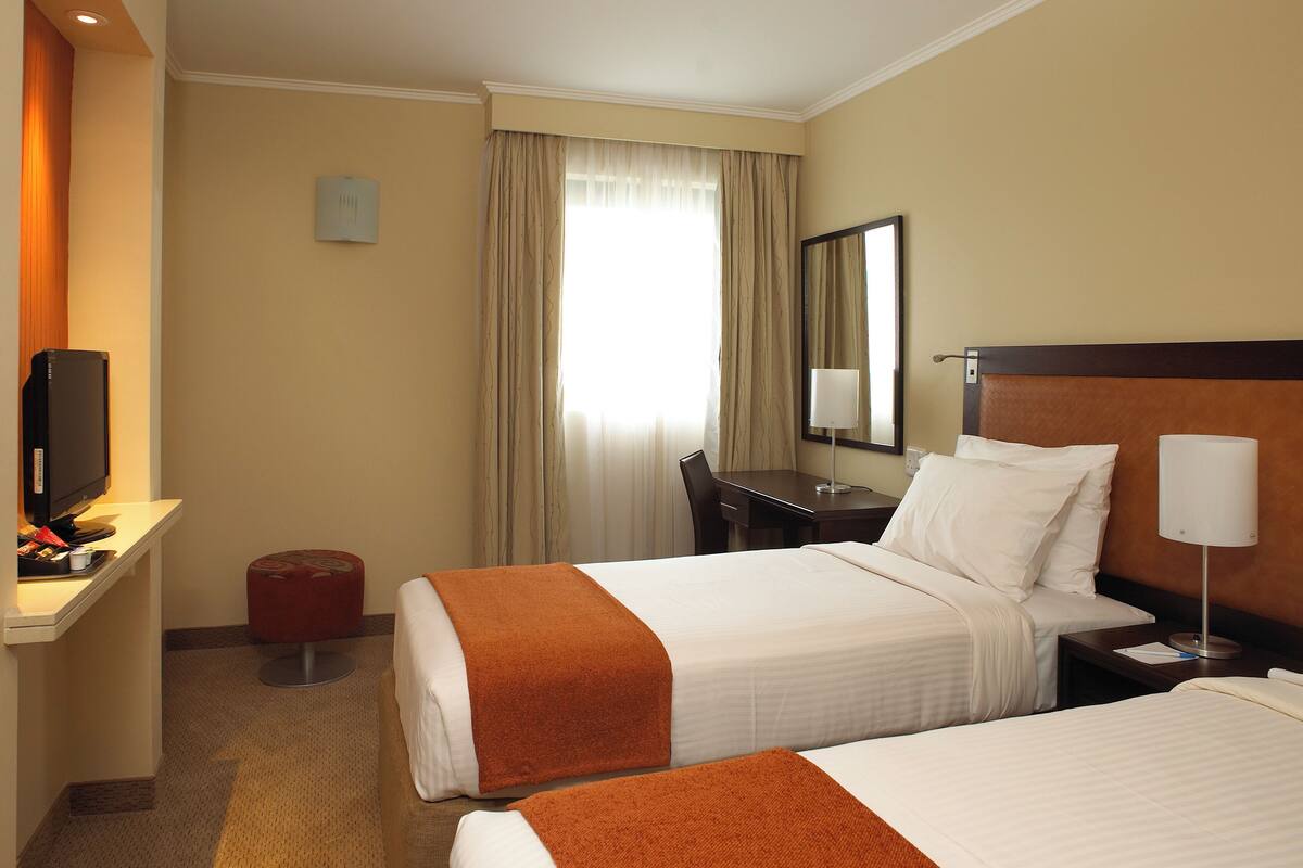 Quarto de hotel com duas camas, uma escrivaninha com computador, uma cadeira, uma televisão e uma janela com cortinas.