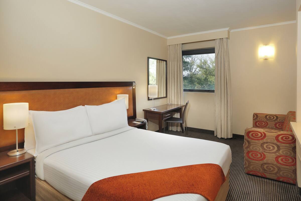Quarto de hotel com cama, escrivaninha, cadeira, televisão e janela com cortinas.